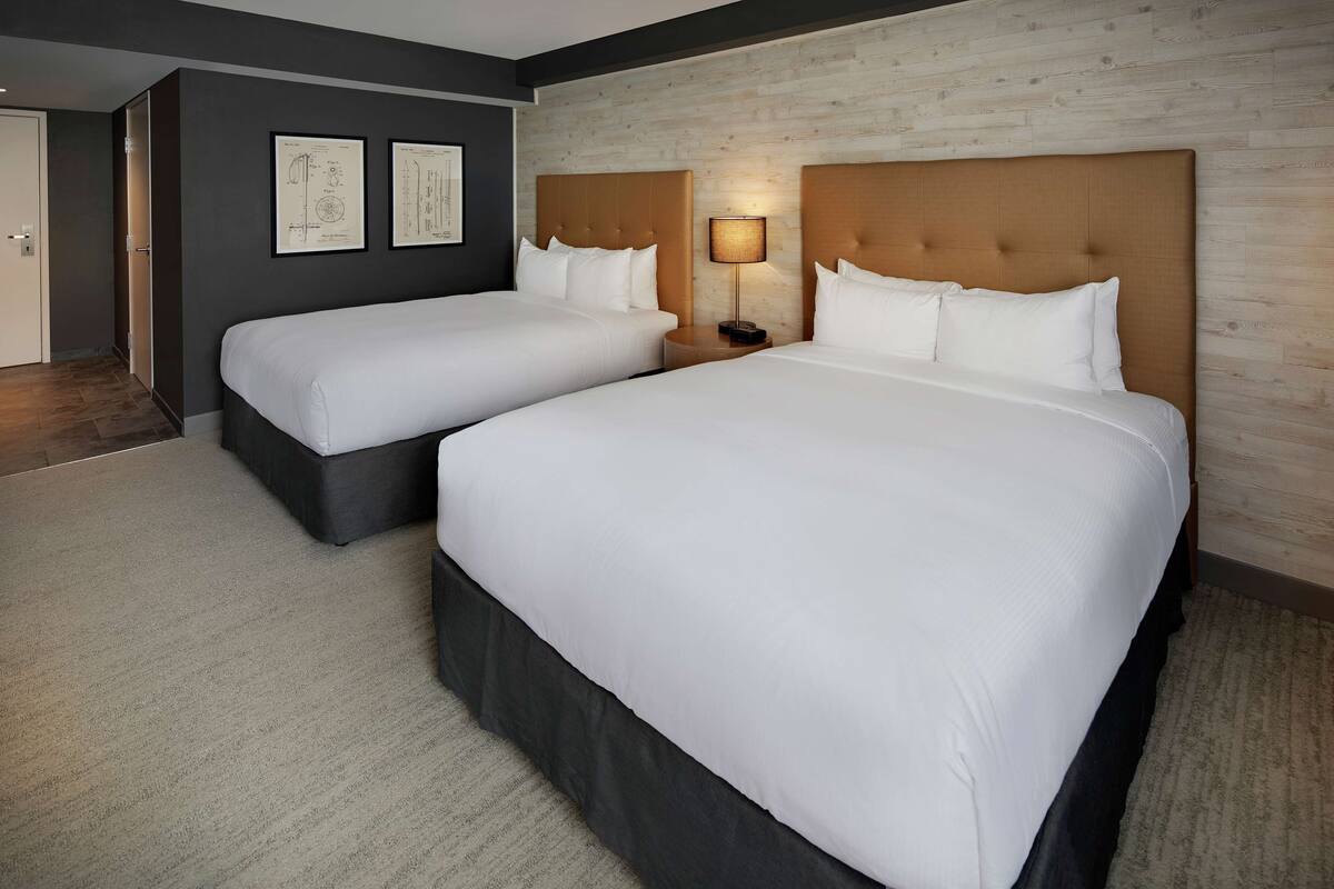 Quarto de hotel com duas camas, cabeceira de madeira, mesa de cabeceira com abajur e diplomas emoldurados na parede.