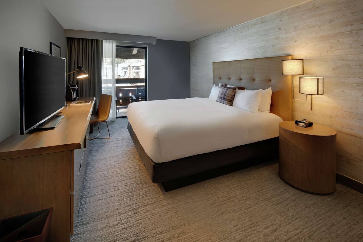 Quarto de hotel com uma cama, uma escrivaninha com televisão, uma cadeira, uma mesinha de cabeceira e vista para o exterior.
