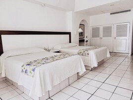 Quarto de hotel com duas camas, roupa de cama branca, cabeceira de madeira, chão de azulejos e guarda-roupa branco com portas venezianas.