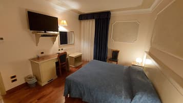Quarto de hotel com cama, escrivaninha, cadeira, televisão e janela com cortinas.
