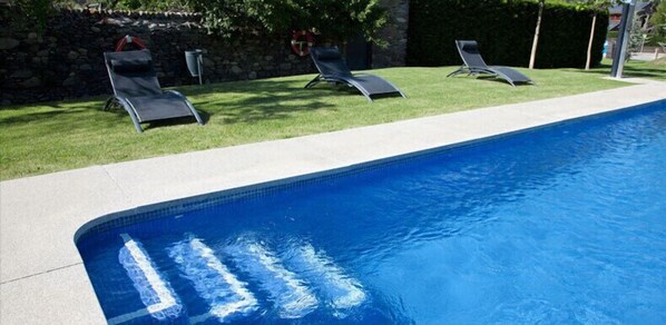 Piscina com água azul cristalina, rodeada por um muro de pedra e uma área gramada com espreguiçadeiras.
