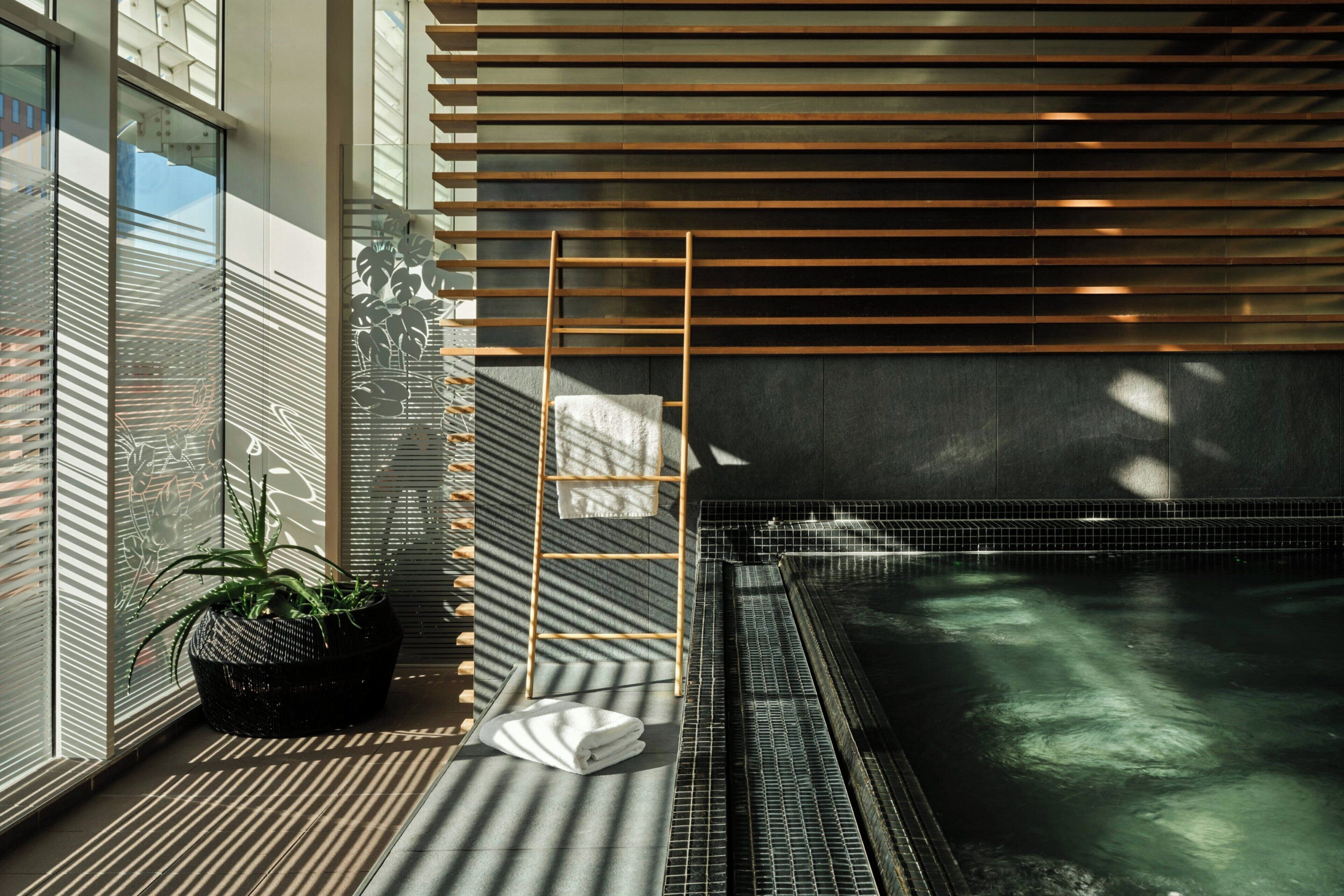 Uma área de piscina interior moderna com teto de ripas de madeira, um pequeno espaço de estar com uma toalha e um vaso de planta.