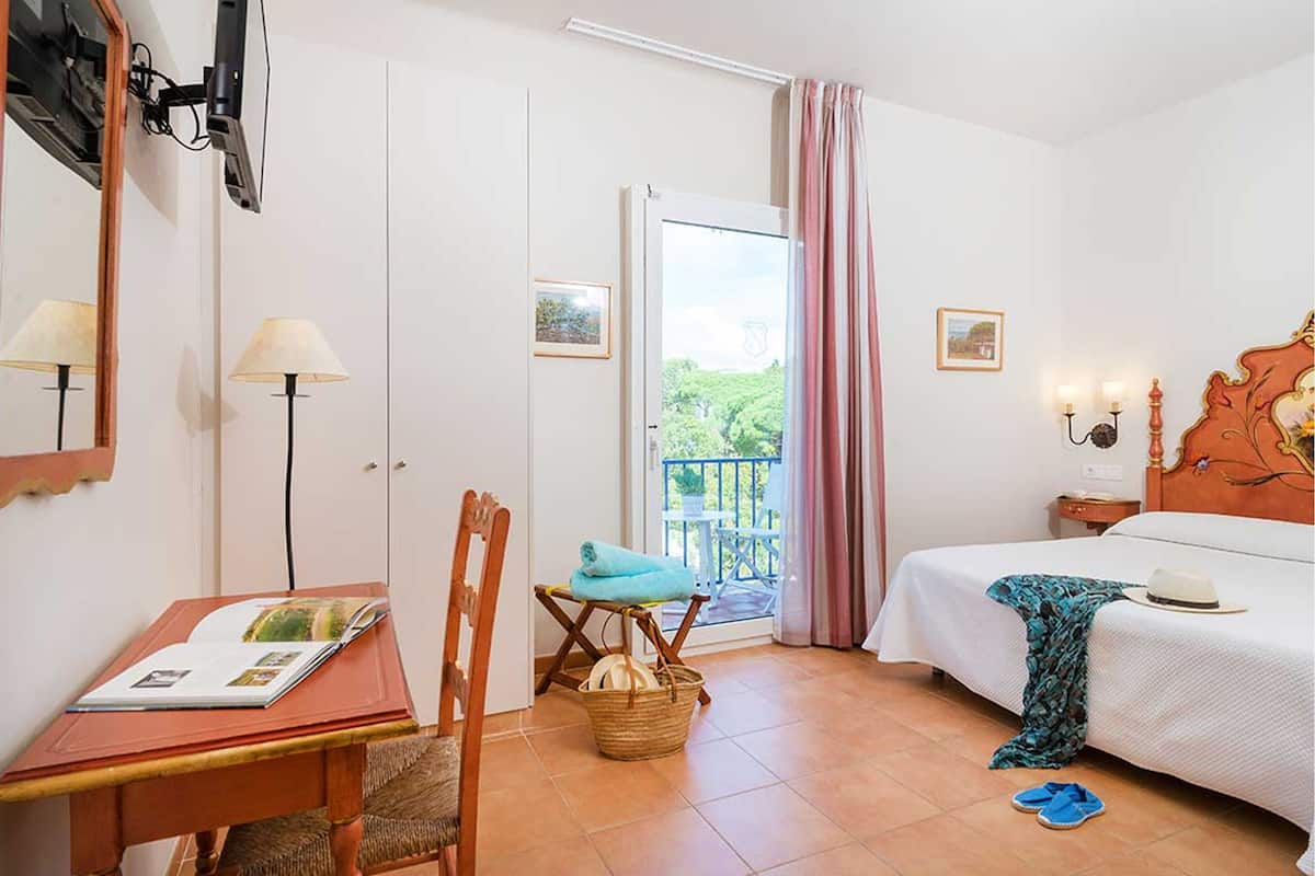 Quarto de hotel com cama, escrivaninha com um livro, cadeira, janela com vista para áreas verdes e varanda.