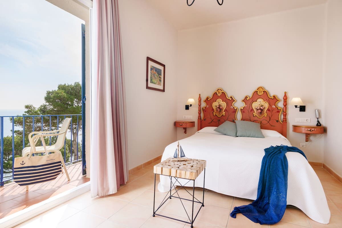 Quarto com cama, mesas de cabeceira, um parapeito com cortinas, varanda com uma cadeira e vista para o mar.