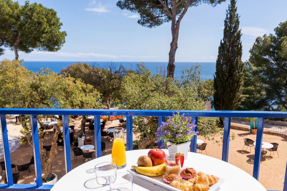 Uma mesa com uma variedade de alimentos para o café da manhã, incluindo frutas, doces e uma bebida, com vista para uma paisagem com mar e árvores.