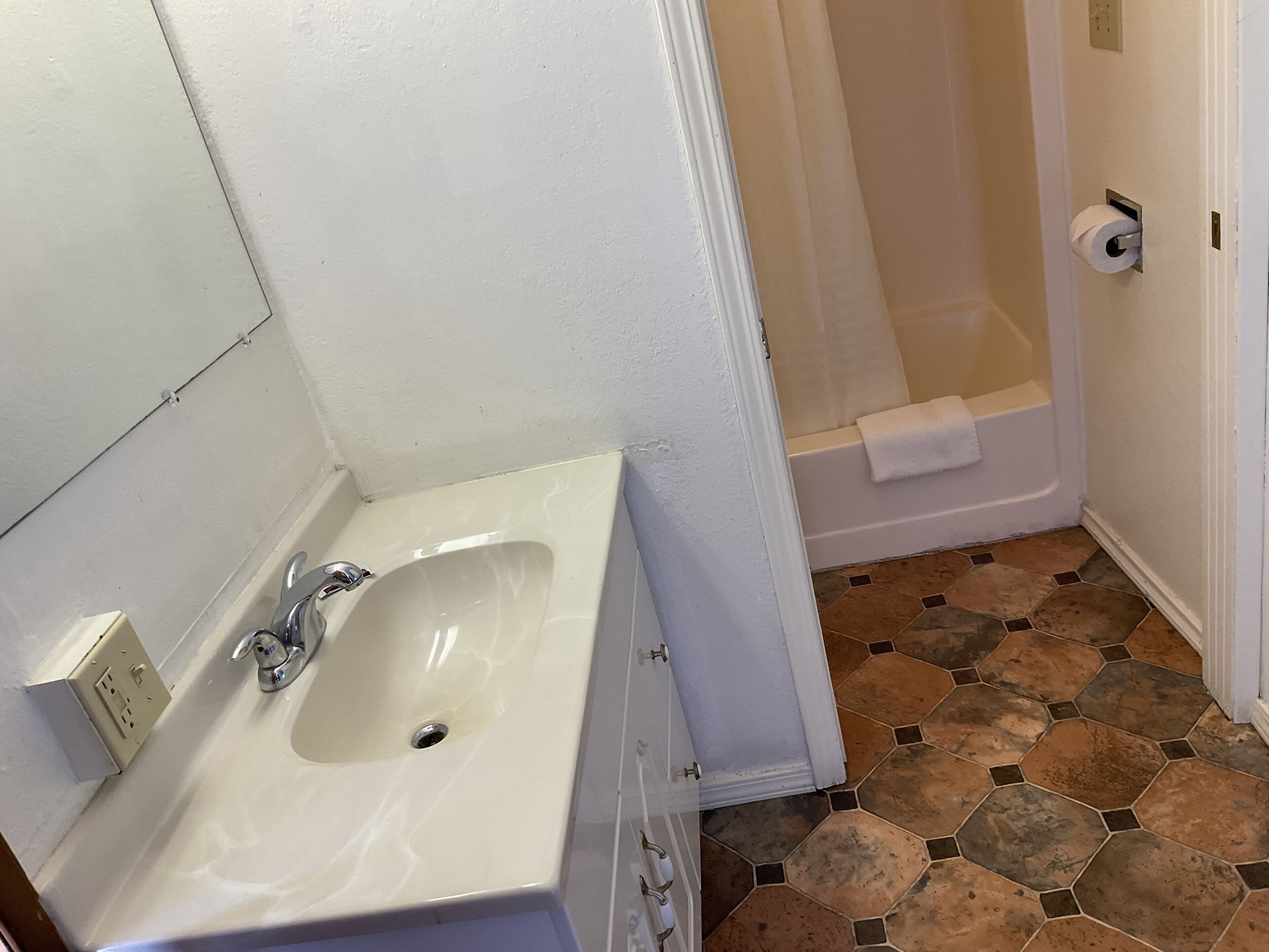 Um banheiro com pia branca, espelho, chuveiro com porta de vidro e porta-papel higiênico.