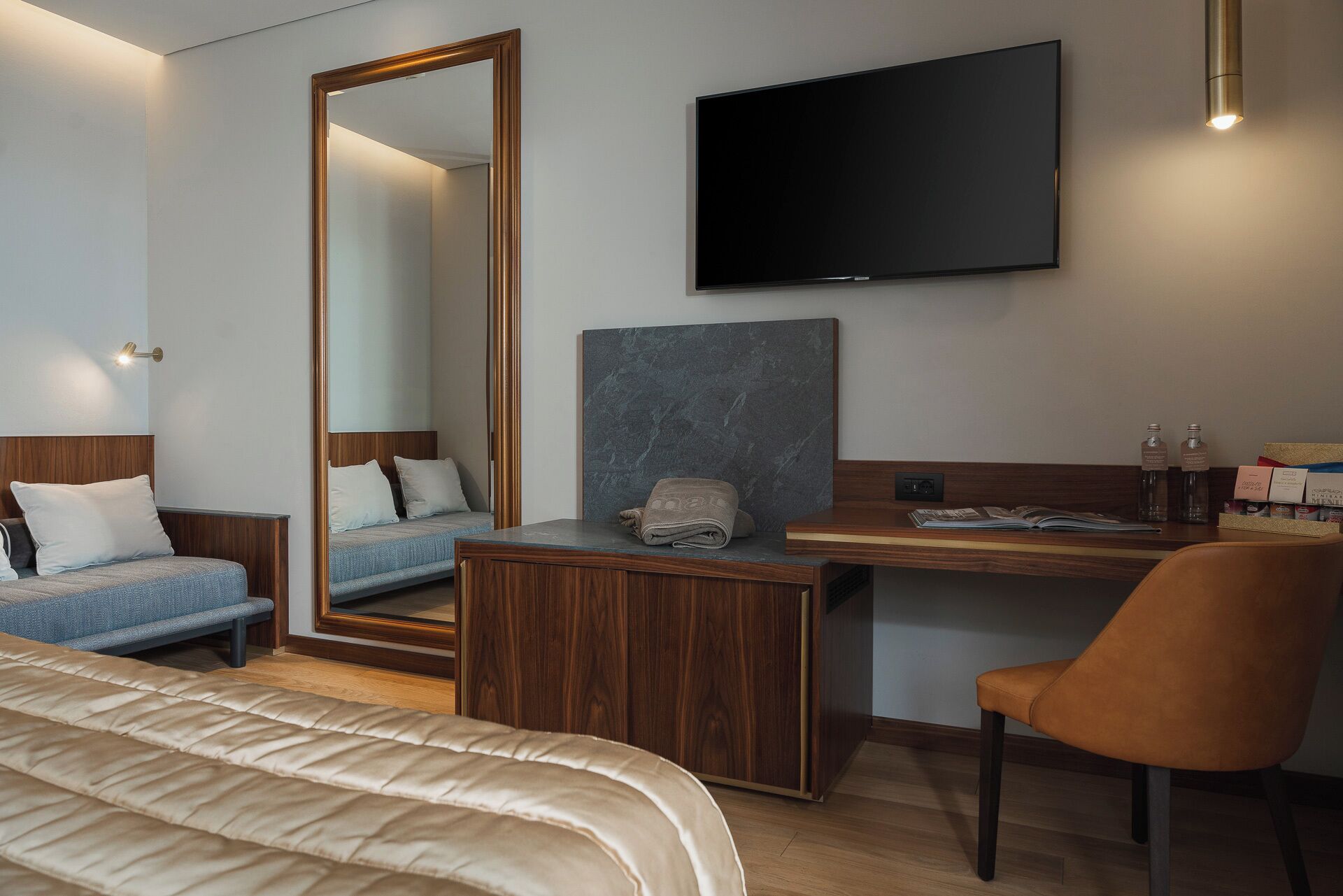 Quarto de hotel com cama, escrivaninha, cadeira, televisão e um espelho grande.