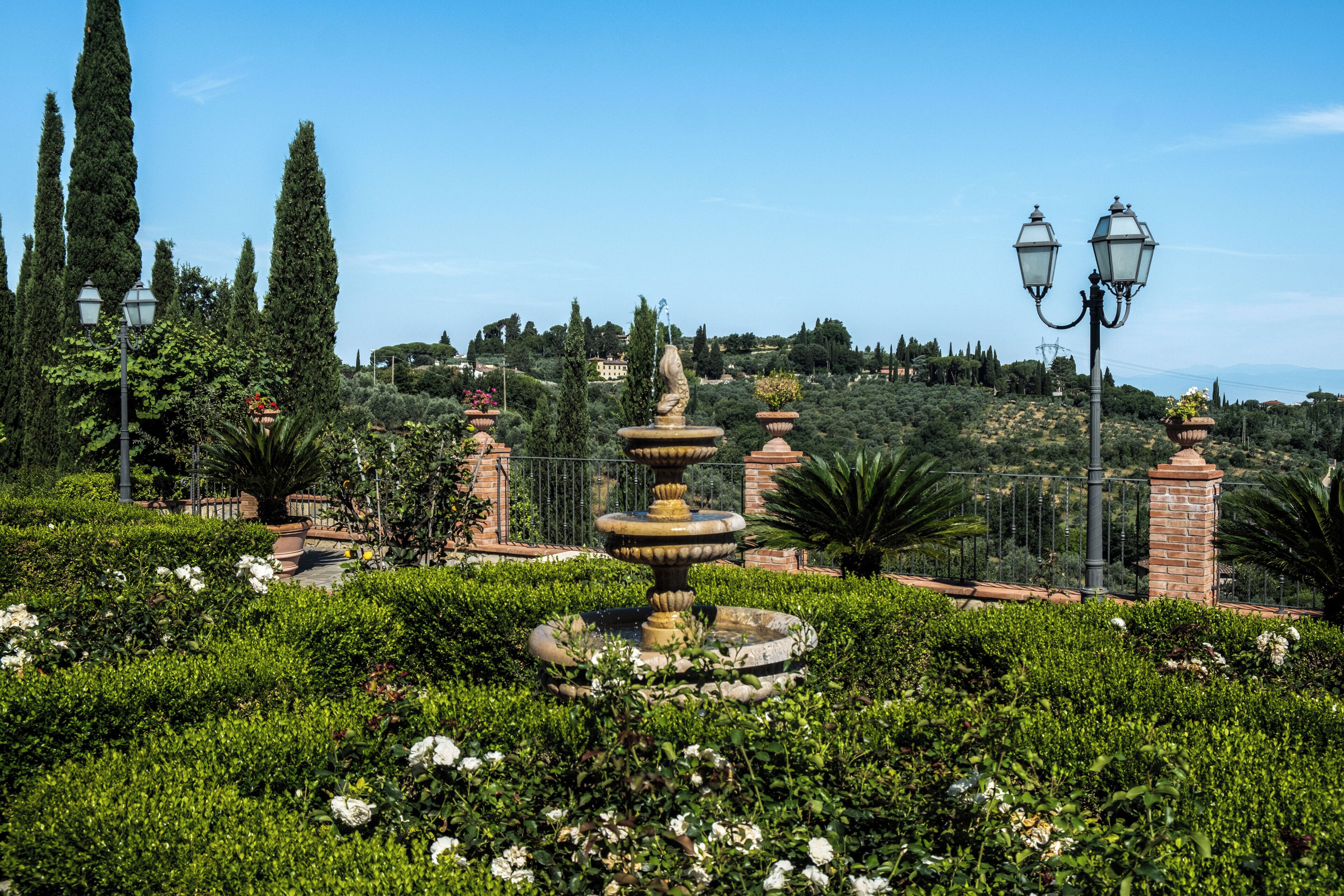 Um jardim com uma fonte, árvores altas e uma cerca.