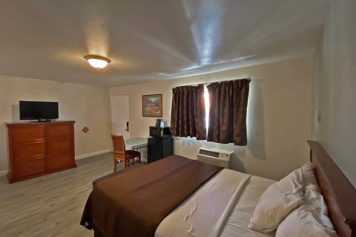 Quarto com cama, penteadeira, escrivaninha, cadeira, televisão, janela com cortinas e luminária no teto.