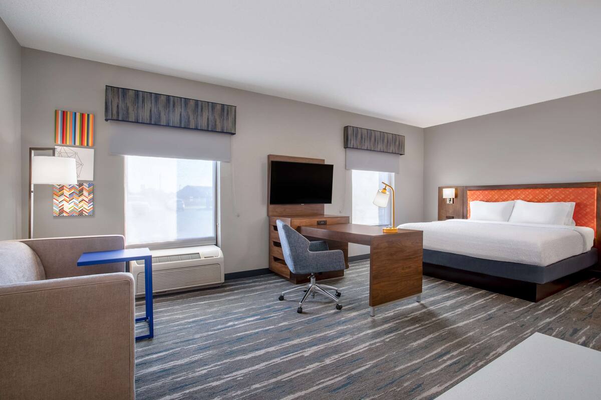 Quarto de hotel com uma cama, escrivaninha, cadeira, televisão e janela.