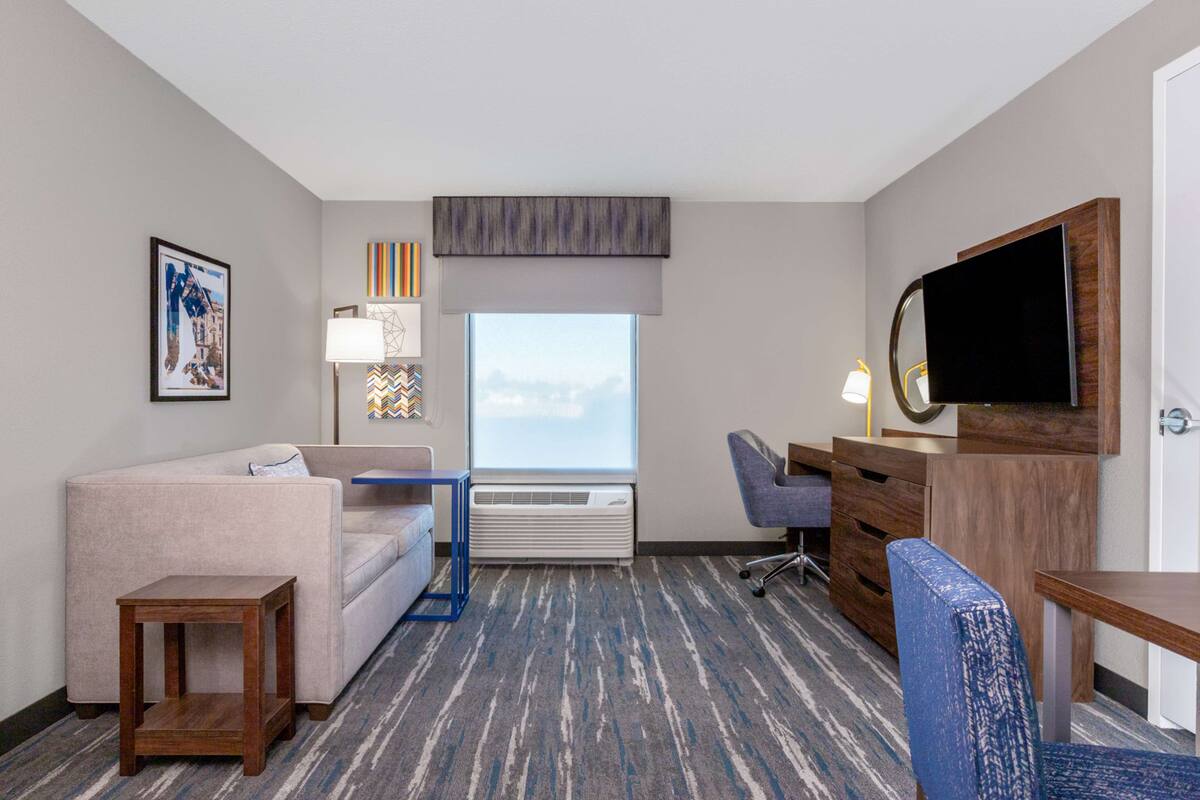 Quarto de hotel com sofá, escrivaninha, cadeira, televisão e janela com vista.