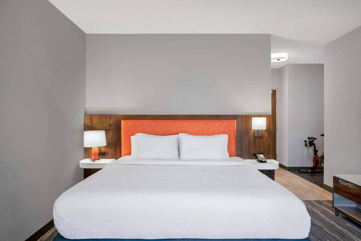 Quarto de hotel com uma cama grande, duas mesas de cabeceira com luminárias, um painel de madeira na cabeceira e uma bicicleta ergométrica no canto.