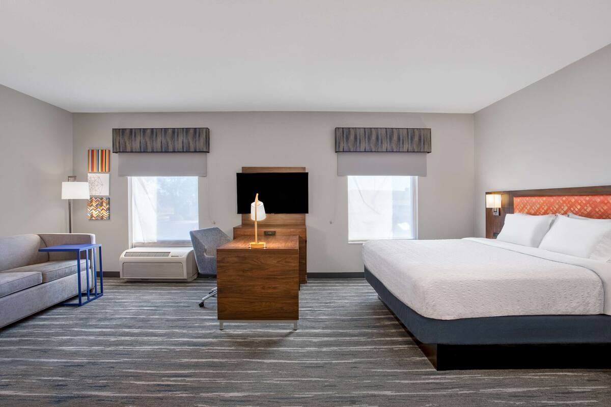 Quarto de hotel com cama, escrivaninha, cadeira, sofá, televisão e janela.