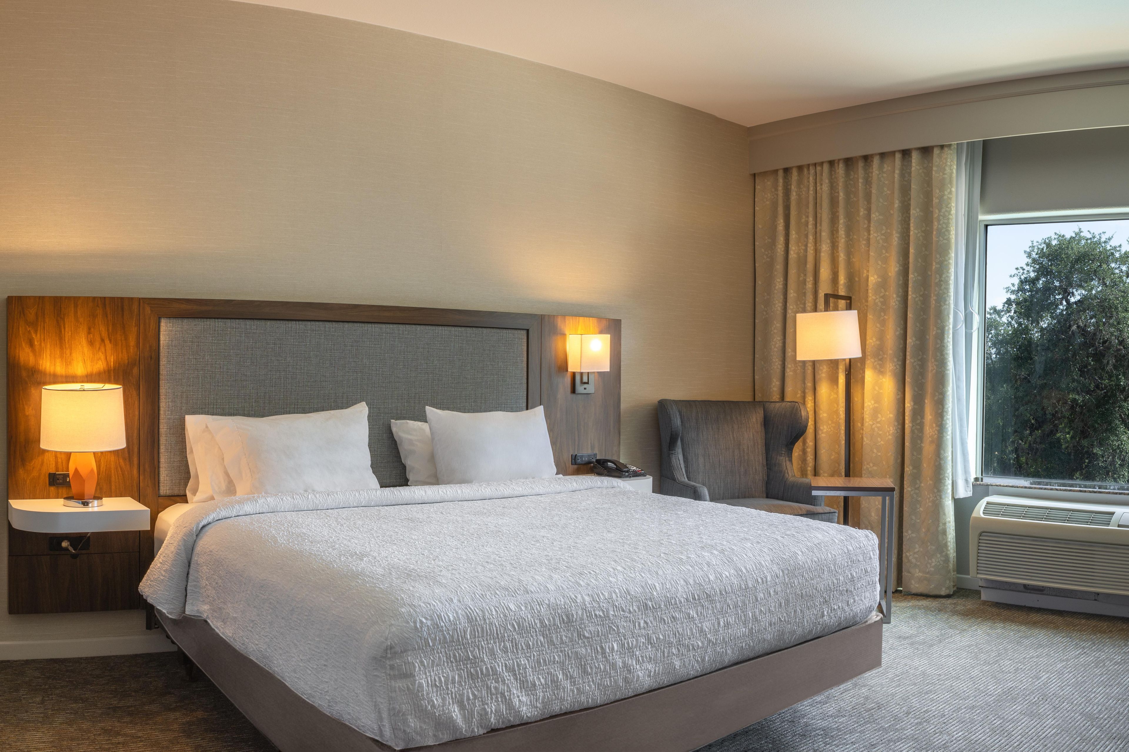 Quarto de hotel com cama grande, duas luminárias de cabeceira, uma cadeira e vista para o exterior.