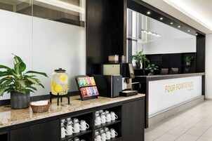 Uma área de recepção com balcão exibindo uma máquina de café, um dispensador de água saborizada com frutas e um suporte colorido para cápsulas de café. Há prateleiras com canecas brancas e uma planta em vaso.