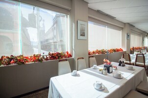 Uma sala de conferências com mesas e cadeiras brancas, e uma janela com flores vermelhas.