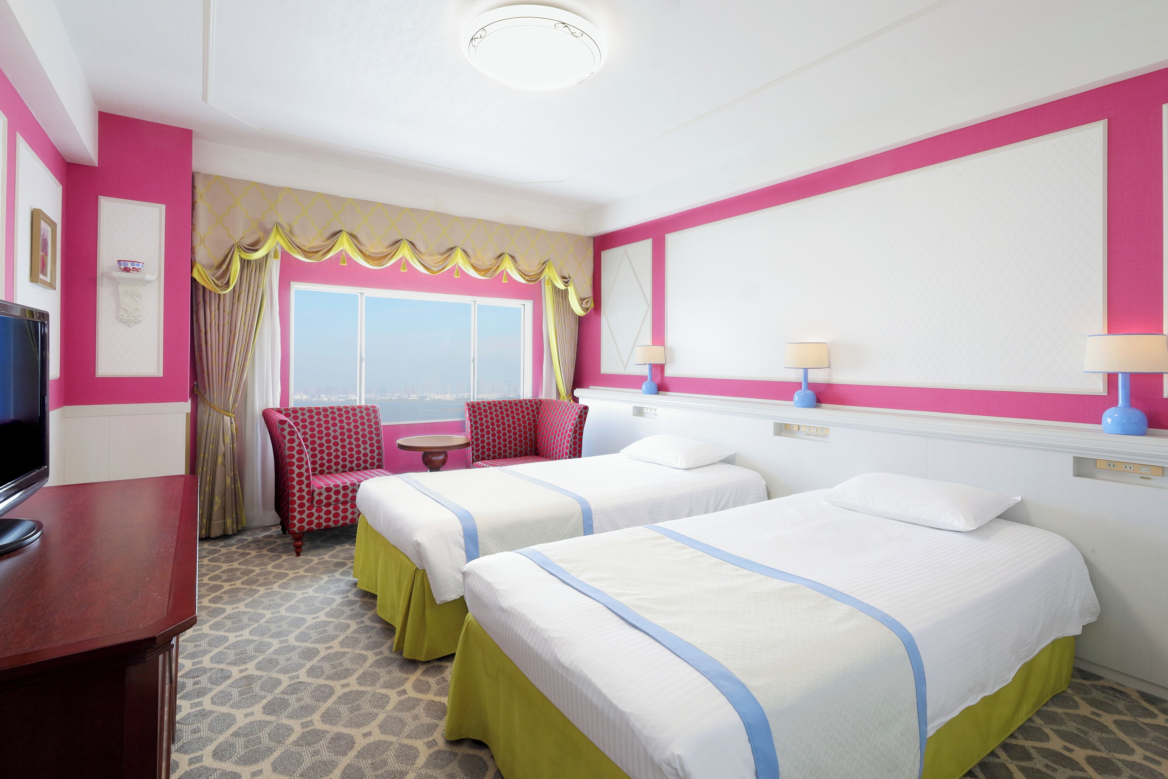 Quarto de hotel com duas camas, uma escrivaninha, televisão, janela com cortinas e vista para a cidade.