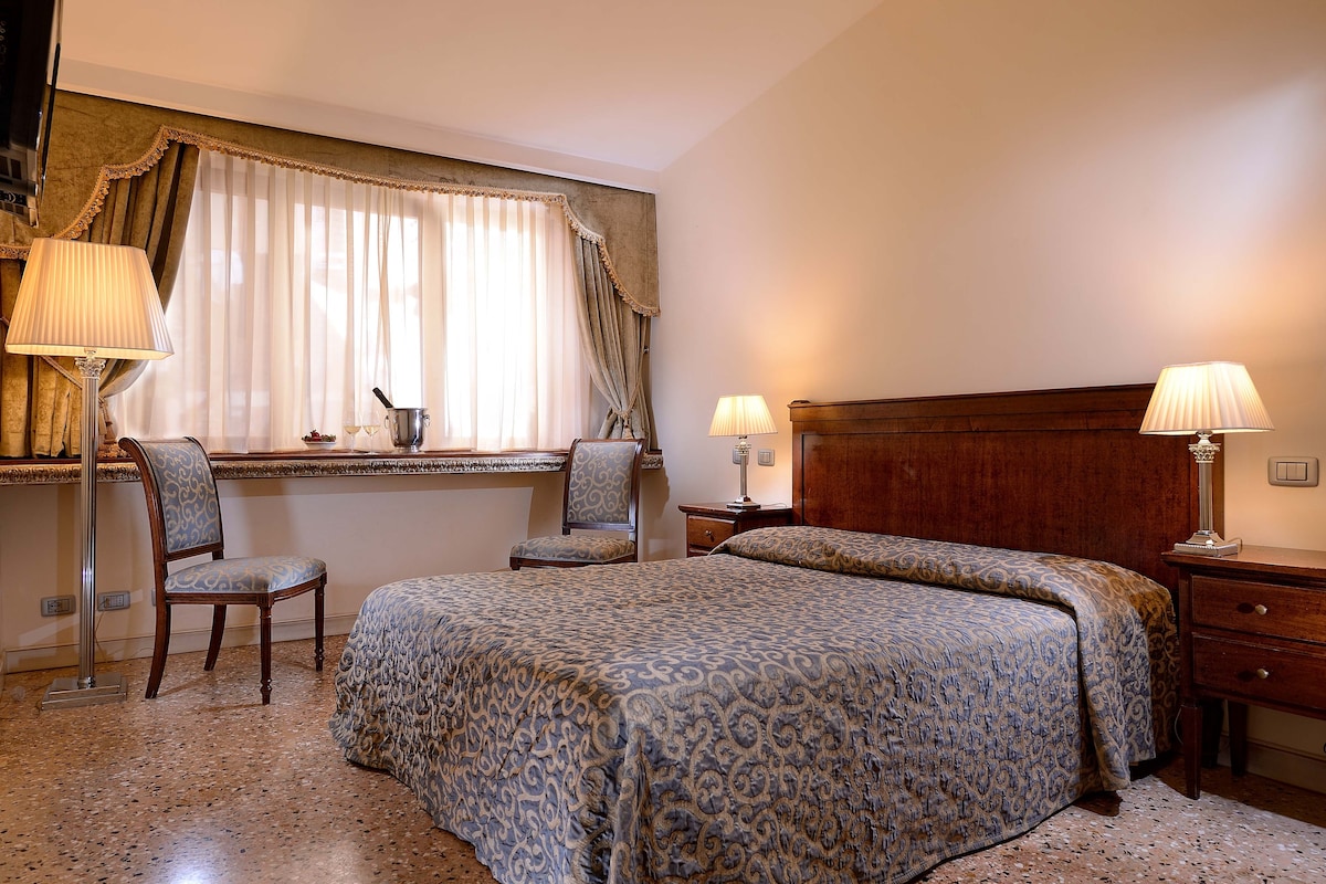 Quarto de hotel com cama, duas cadeiras, escrivaninha, luminária e janela com cortinas.