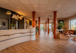 Um saguão de hotel com balcão de recepção, piso de madeira, pilares grandes e plantas de interior.