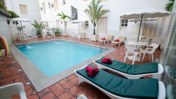 Piscina com espreguiçadeiras e guarda-sóis, rodeada por plantas em vasos e um terraço com ladrilhos.