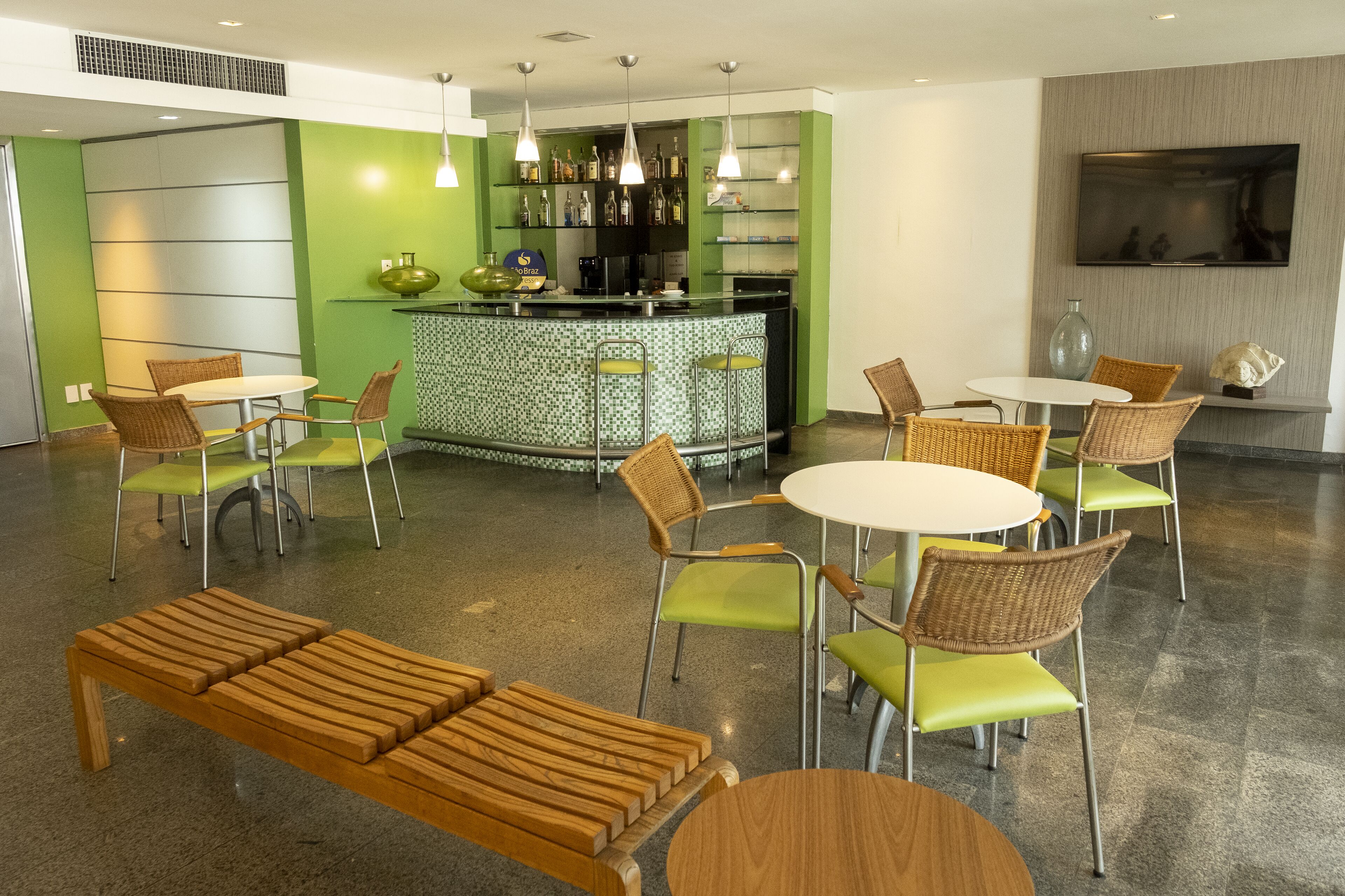 Uma área de bar moderna com paredes verdes, balcão revestido de azulejos e diversas opções de assento, incluindo mesas redondas e cadeiras de vime.