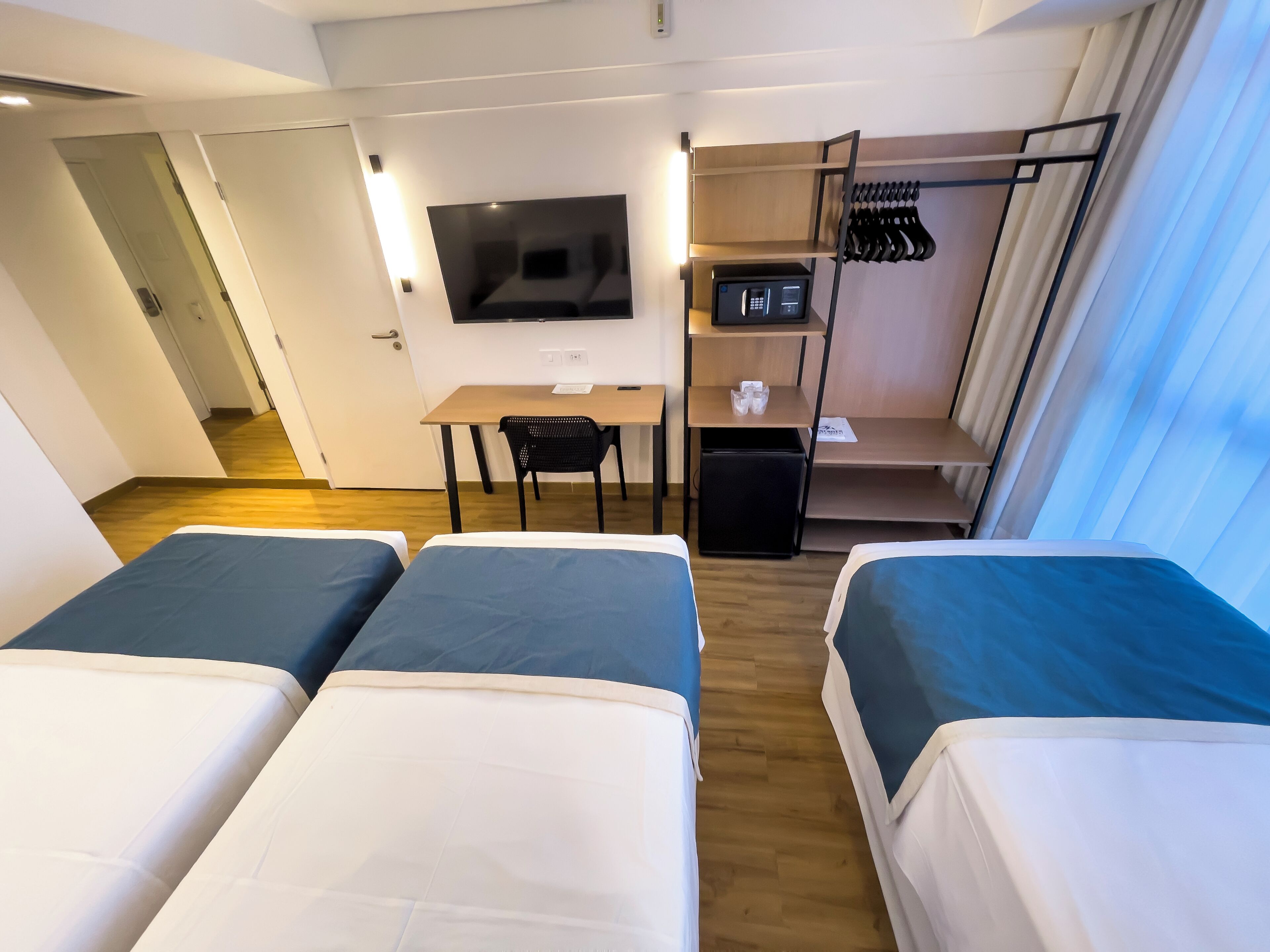 Quarto de hotel com duas camas, uma escrivaninha, televisão, guarda-roupa e uma pequena cozinha equipada.