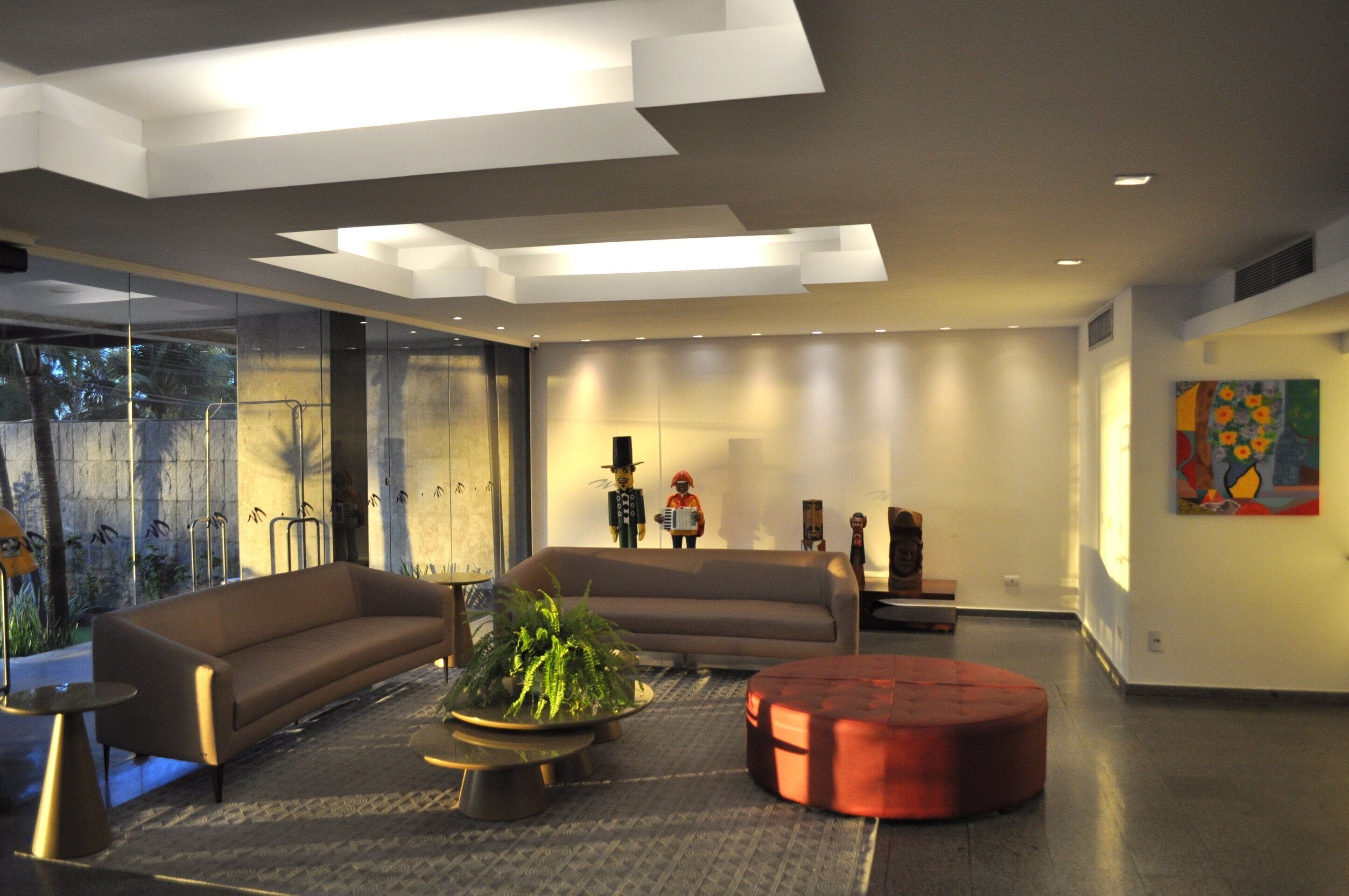 Uma sala de estar moderna com sofá, mesa de centro e um banquinho, parede de vidro com vista e um quadro na parede.