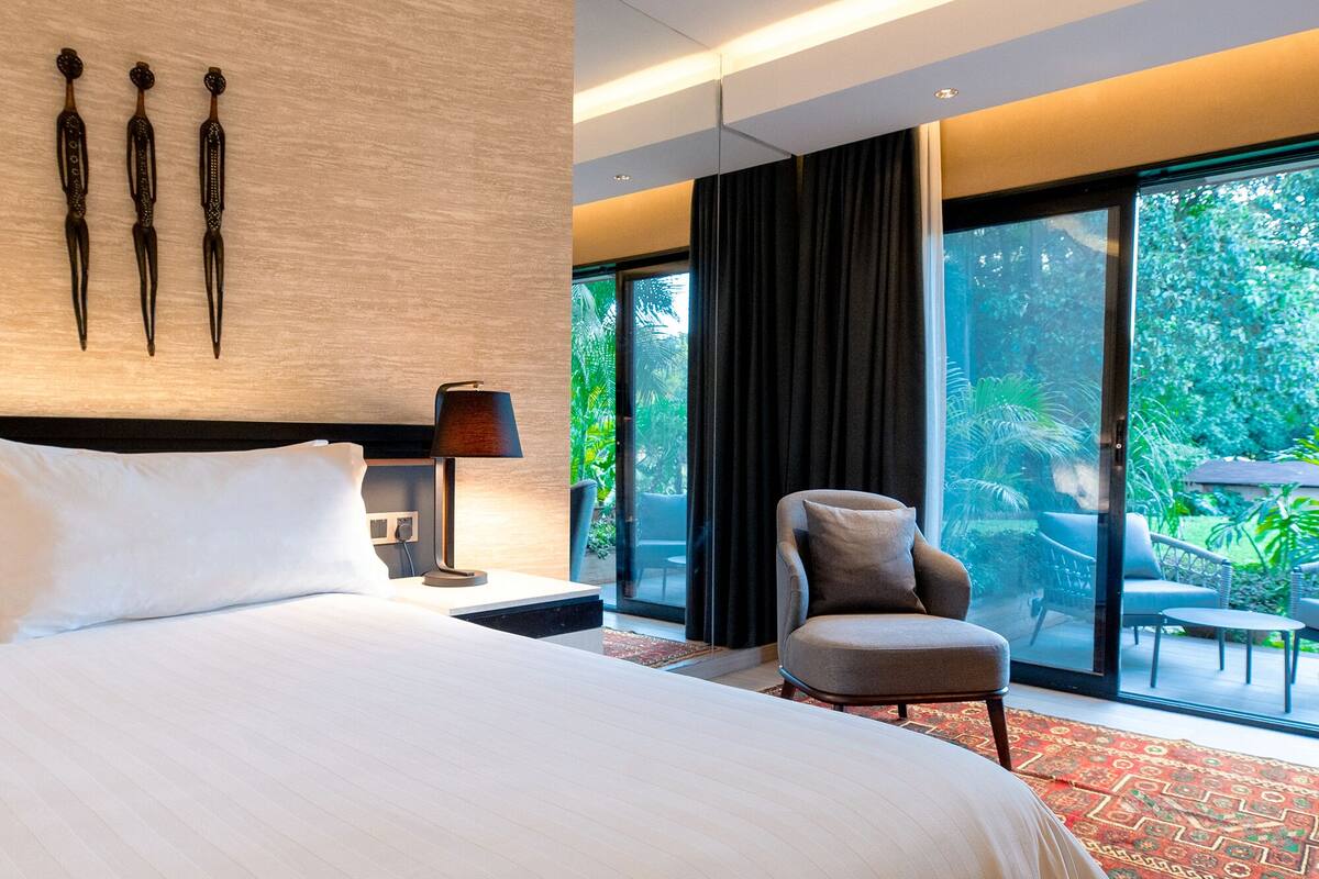 Quarto de hotel moderno com uma cama grande, mesa de cabeceira, luminária, cadeira e vista para o exterior através de uma porta de vidro.