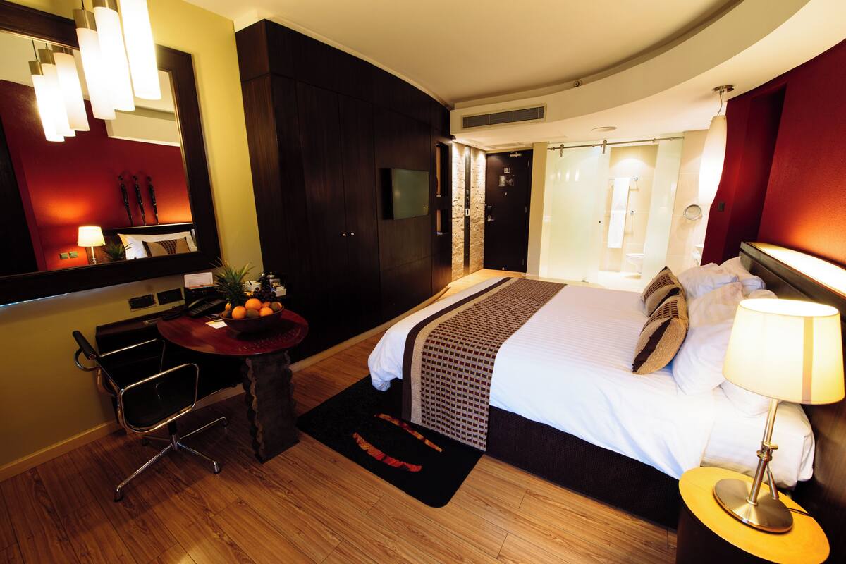 Quarto de hotel com cama, mesa de cabeceira, luminária, escrivaninha com cadeira e espelho.