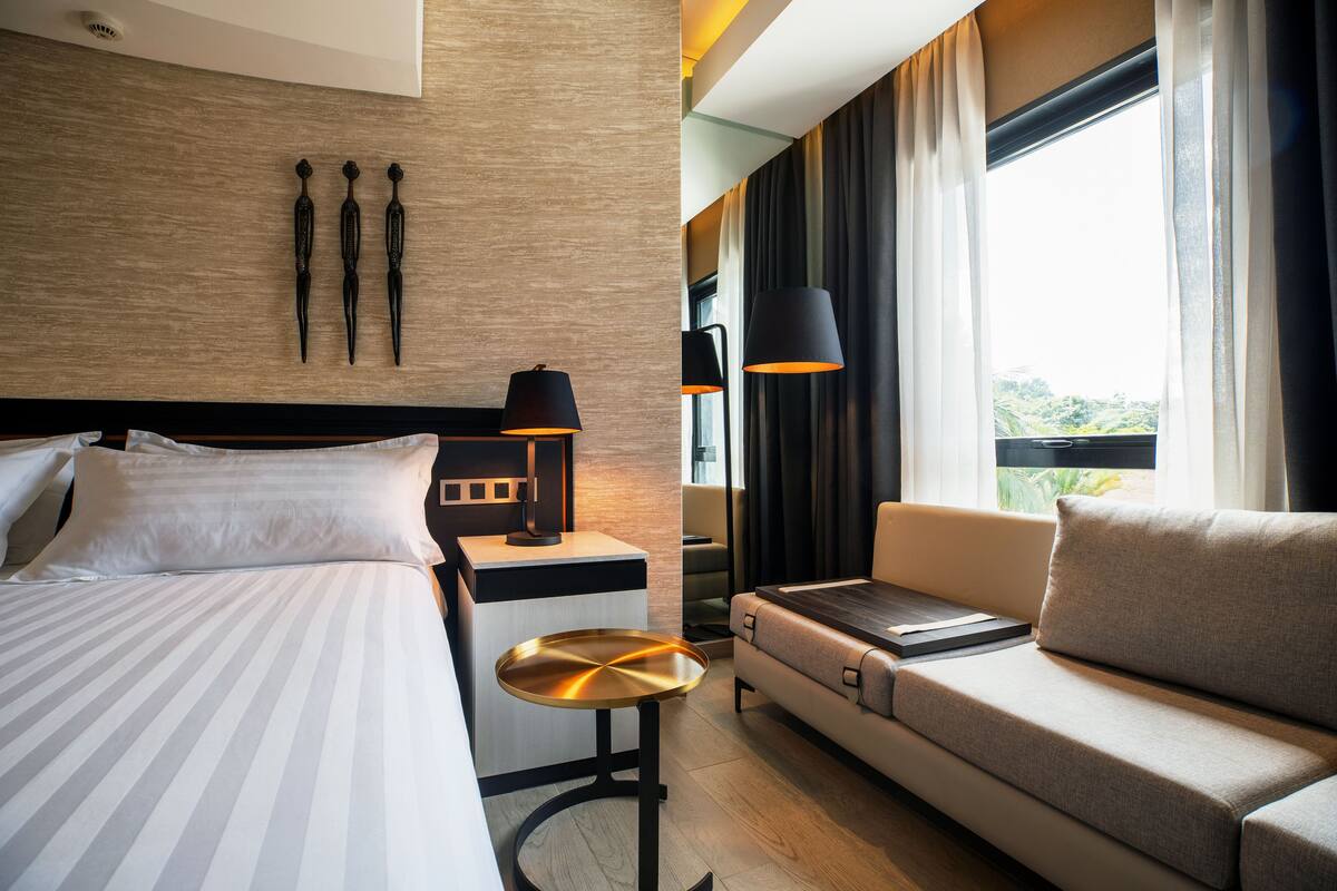 Quarto de hotel com cama, sofá, mesa pequena e mesa de cabeceira. Há luminárias na parede e uma janela com cortinas.
