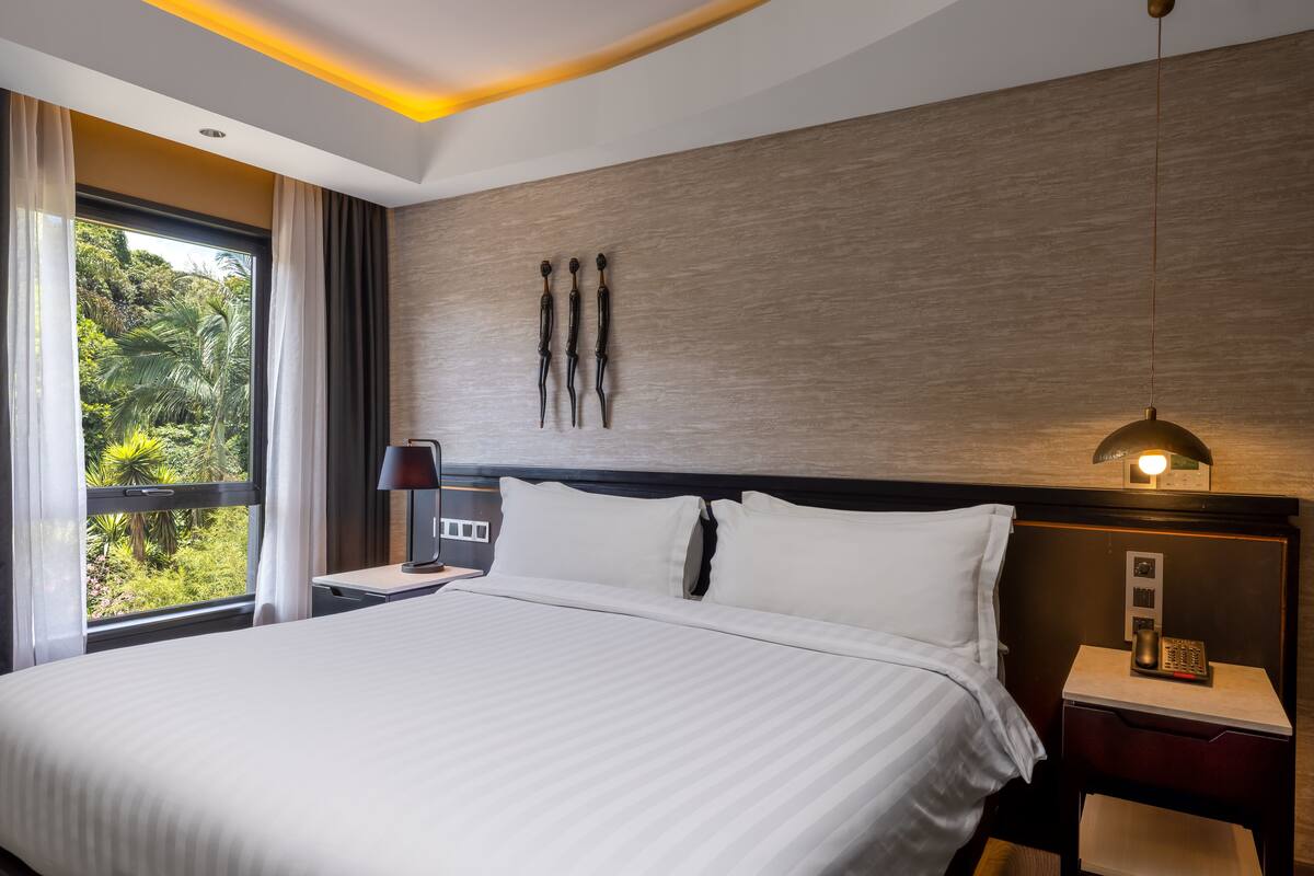 Quarto de hotel moderno com uma cama grande, uma mesa de cabeceira com abajur e vista para o verde lá fora.
