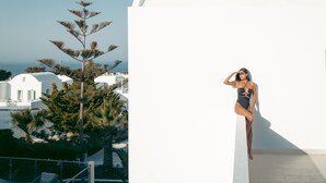 Mulher de biquíni apoiada em parede branca, com vista para edifícios brancos e palmeiras ao fundo.