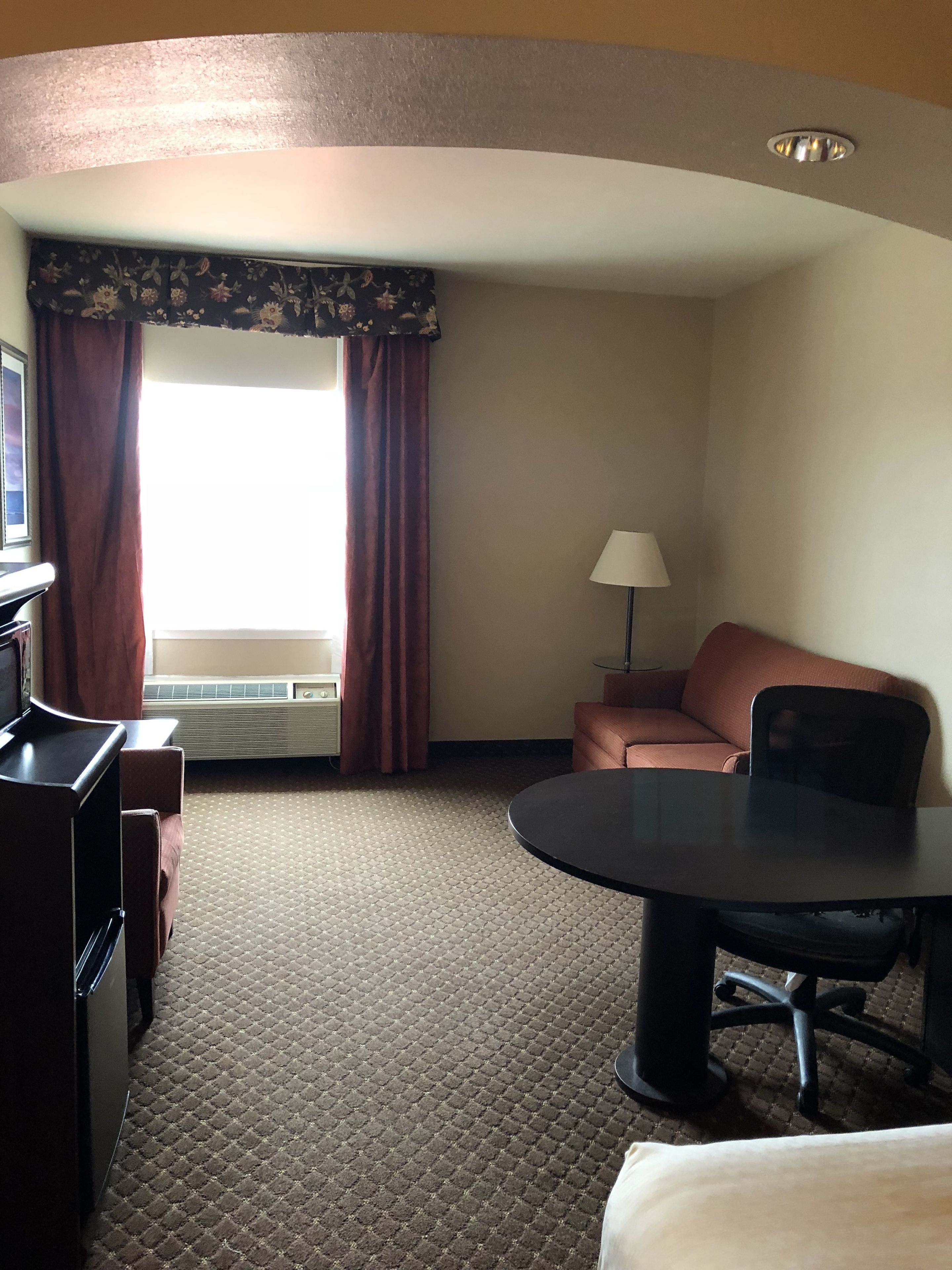 Quarto de hotel com cama, escrivaninha, cadeira, mesa, abajur e janela com cortinas.