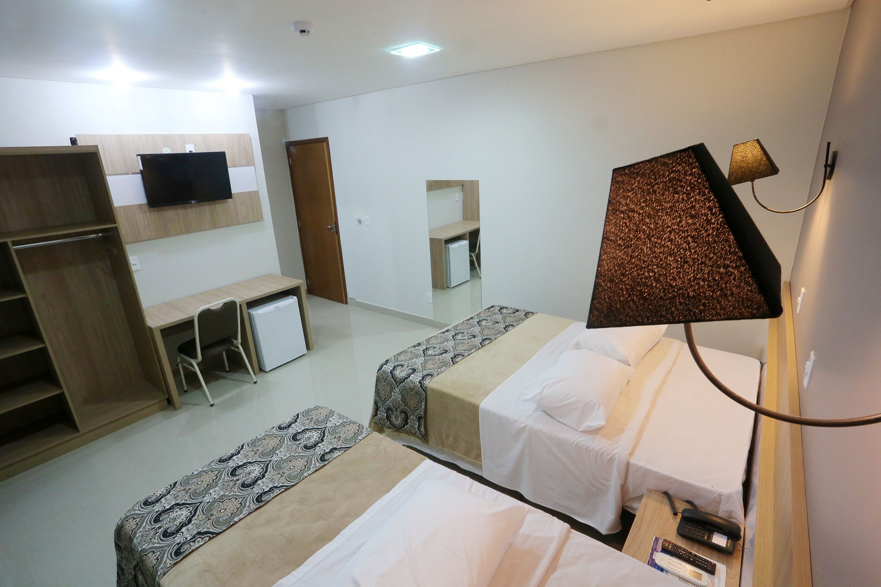 Quarto de hotel com duas camas, uma escrivaninha, uma TV e uma luminária.