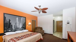 Quarto de hotel com cama, uma cadeira, ventilador de teto e um pôster com vista para a cidade na parede.