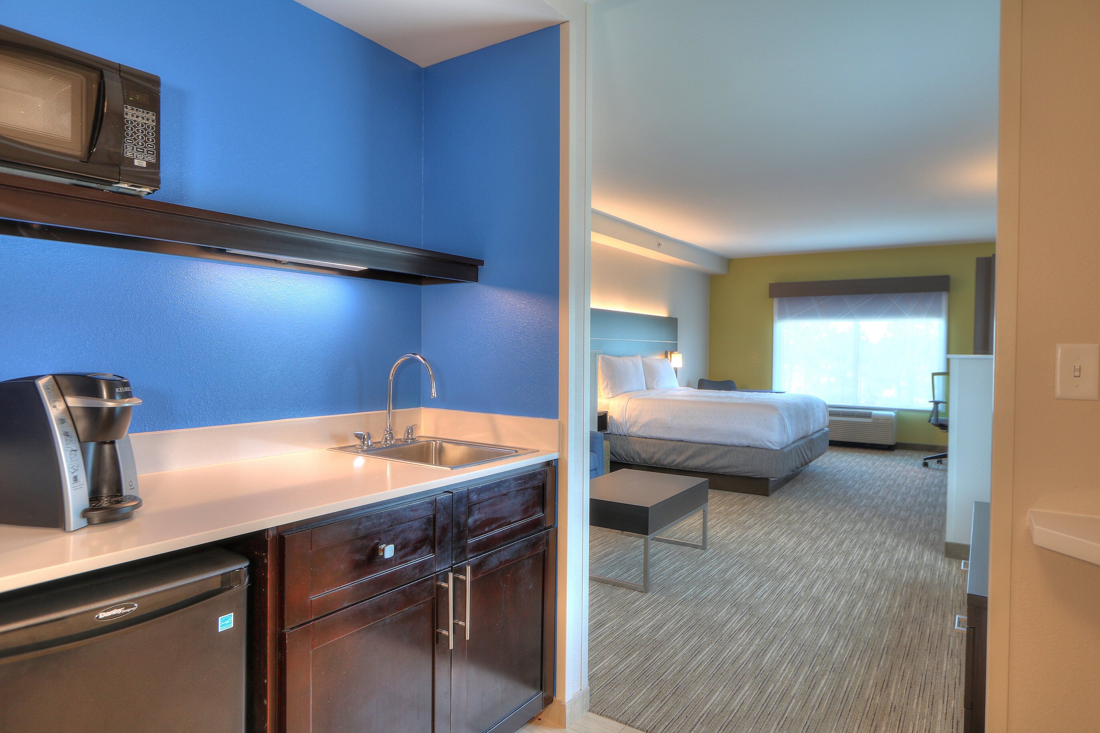 Quarto de hotel com cozinha compacta, cama, mesa de cabeceira e janela.