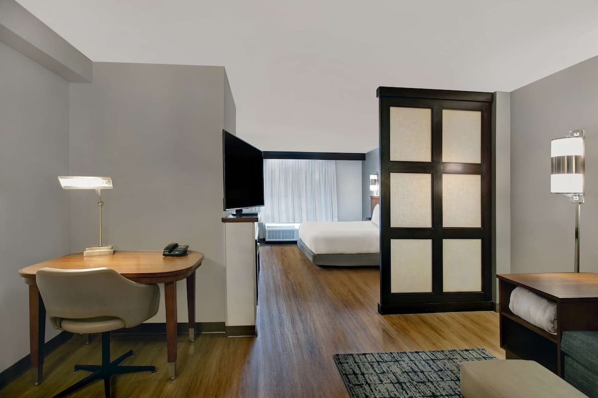 Quarto de hotel com escrivaninha, cadeira, cama e uma janela grande com cortinas.