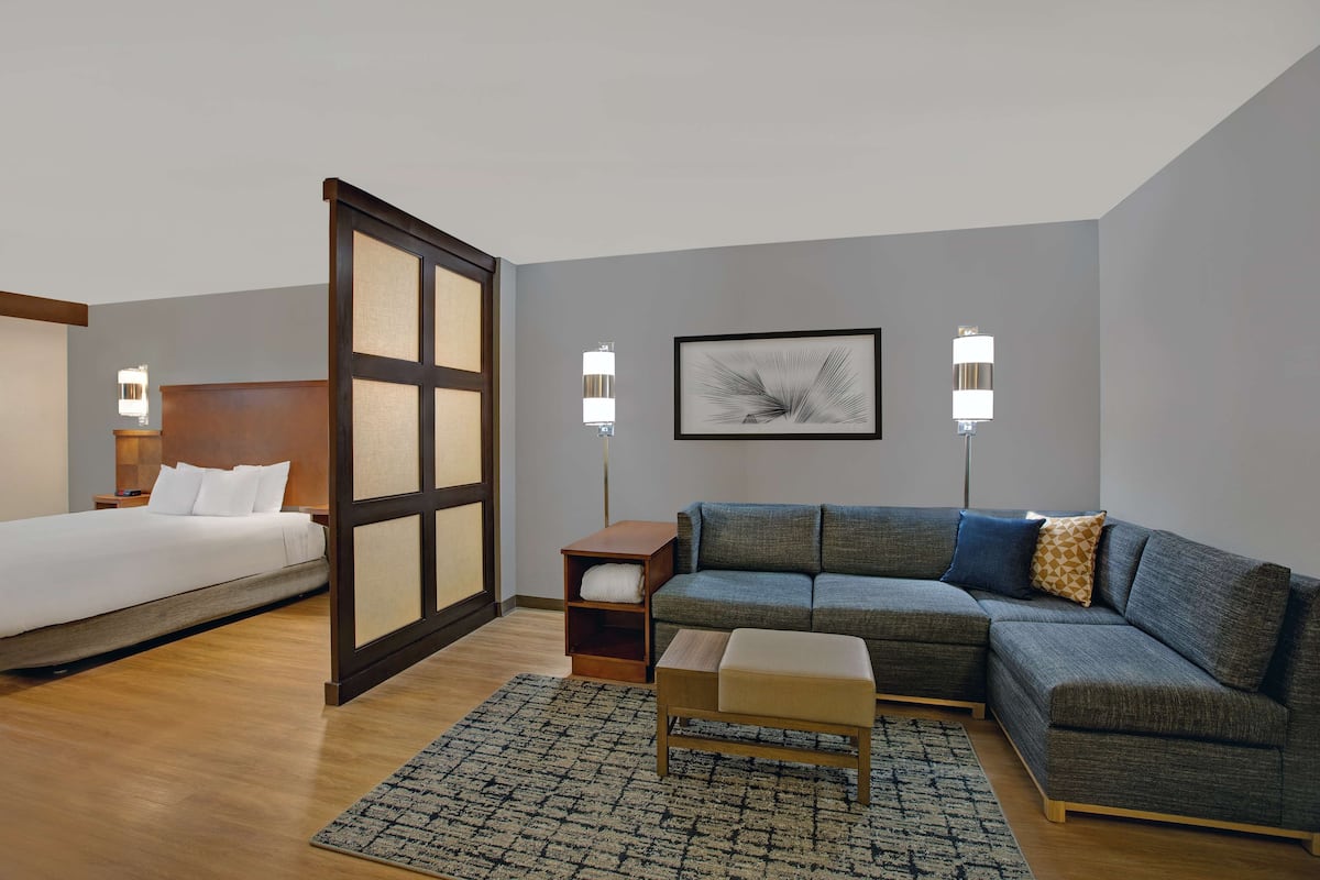 Quarto de hotel com cama, sofá, mesinha, abajur e quadro na parede.