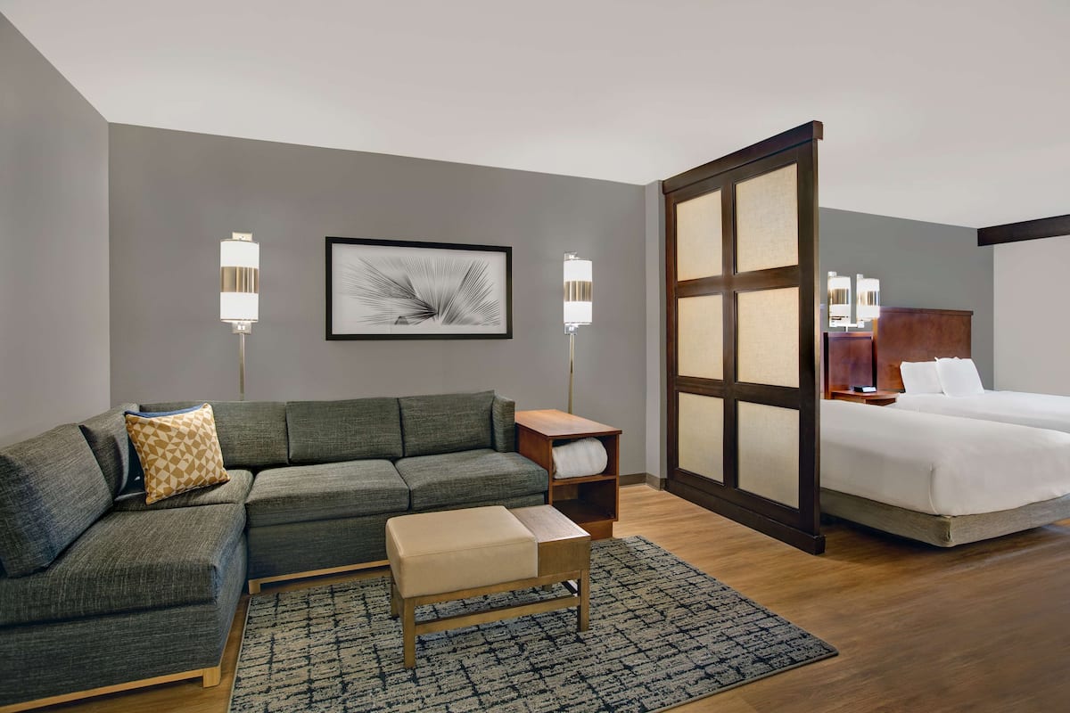Quarto de hotel moderno com sofá cinza, um pequeno banco de madeira, tapete estampado, cama com roupa de cama branca e duas luminárias fixadas na parede.
