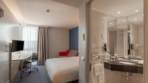 Quarto de hotel com cama, escrivaninha, cadeira, televisão e banheiro com pia e espelho.
