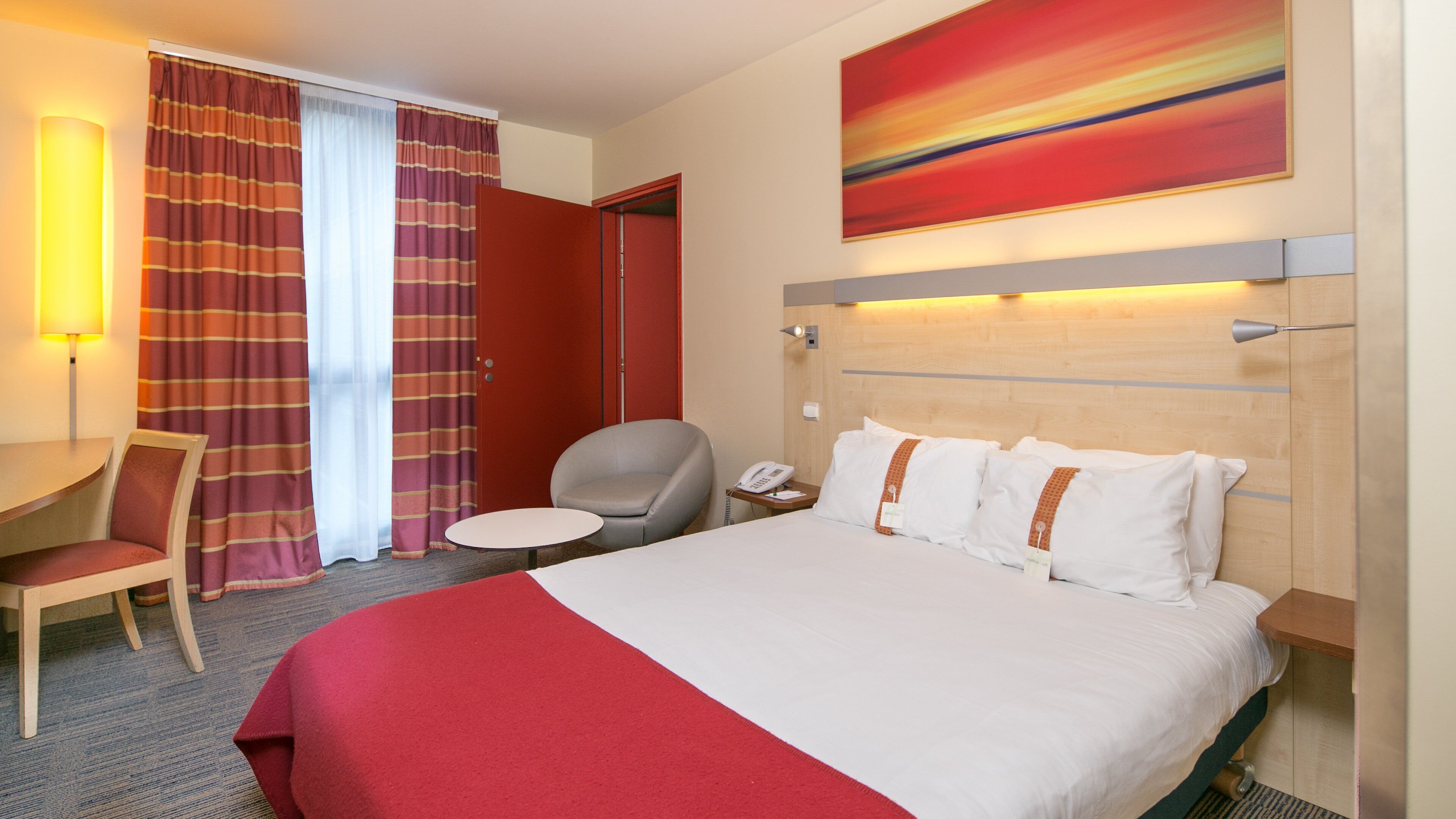 Quarto de hotel com cama, escrivaninha, cadeira, telefone e um quadro na parede.
