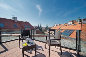 Um terraço no telhado com duas cadeiras de vime, uma mesa pequena com um vaso de flores amarelas e uma vista desobstruída do horizonte da cidade.
