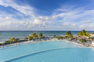 Uma piscina ampla e translúcida em azul, com vista para o oceano e palmeiras.