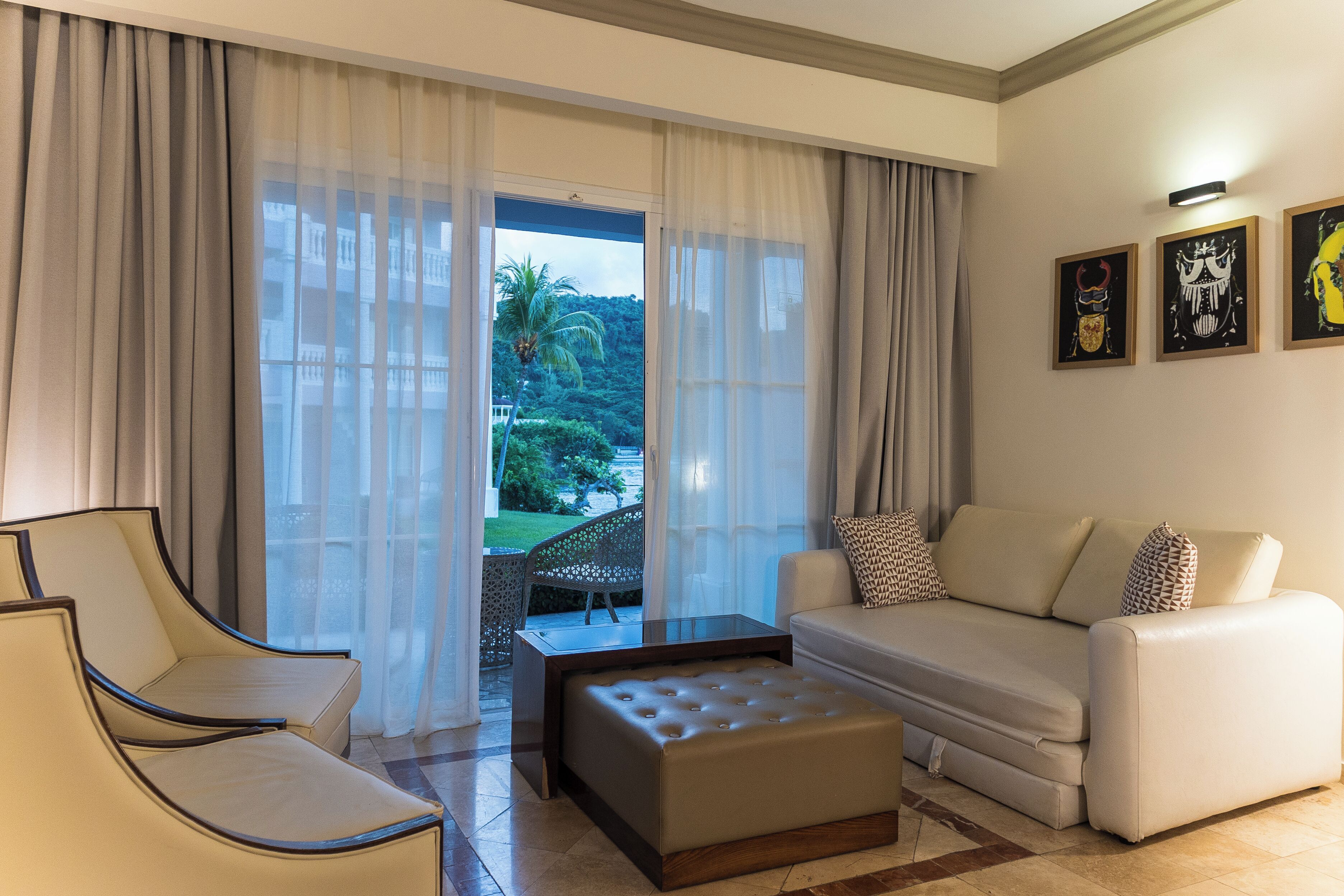 Uma sala de estar moderna com sofá, banqueta e poltrona, uma mesa de centro de vidro e vista para uma varanda com uma palmeira.