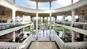 Um amplo átrio com vários níveis, uma escadaria central, colunas brancas e vista para um jardim exuberante.
