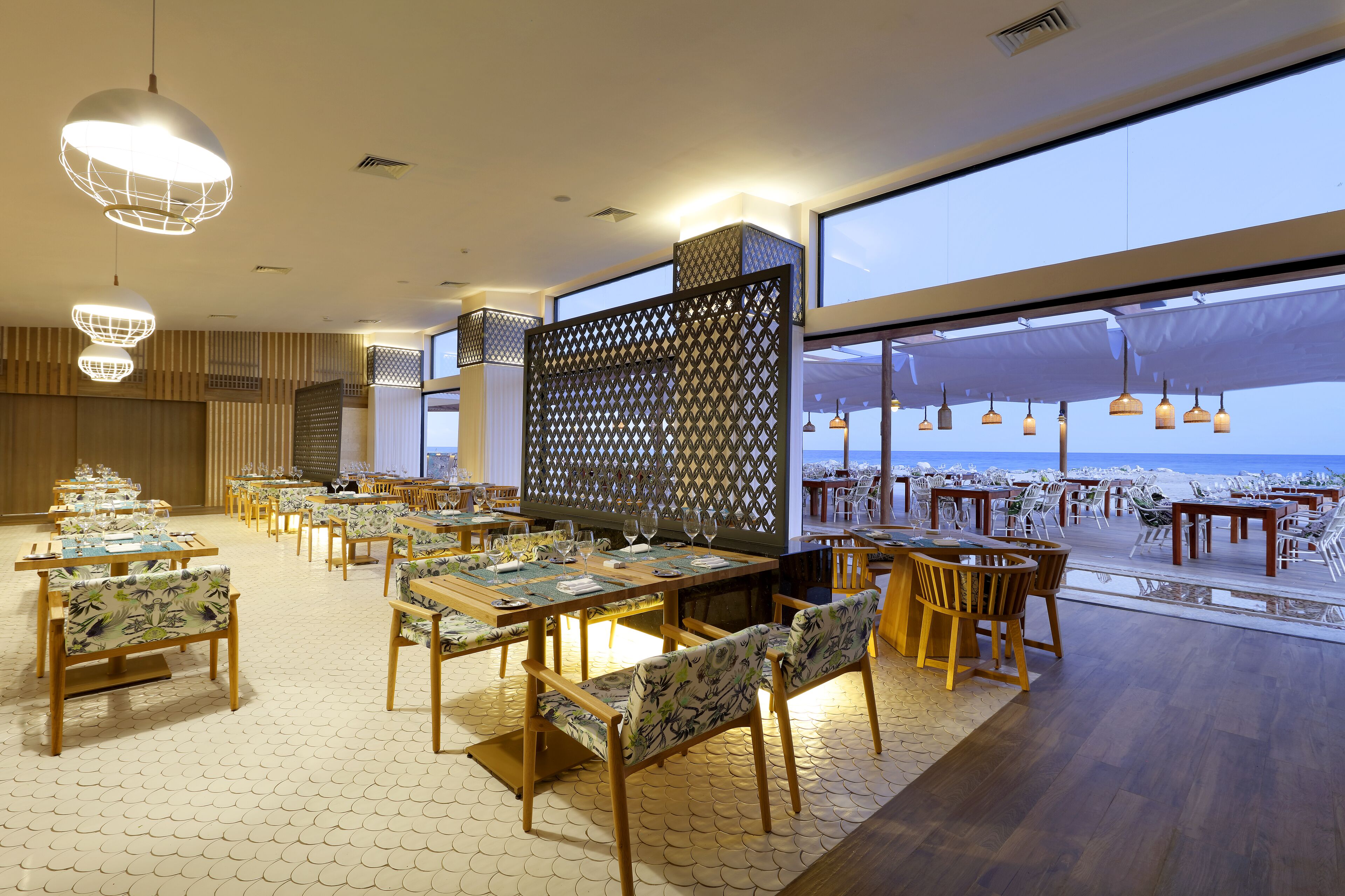 Um restaurante com mesas e cadeiras, uma parede decorativa e grandes janelas com vista para o oceano.