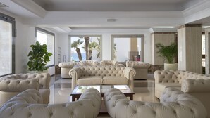 Uma sala de estar espaçosa com um amplo sofá em formato de L, uma mesa de centro e várias poltronas. Há grandes janelas com vista para palmeiras e um edifício.