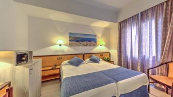 Quarto de hotel com cama, abajures, pintura de uma praia, mesa de cabeceira de madeira e uma cadeira.
