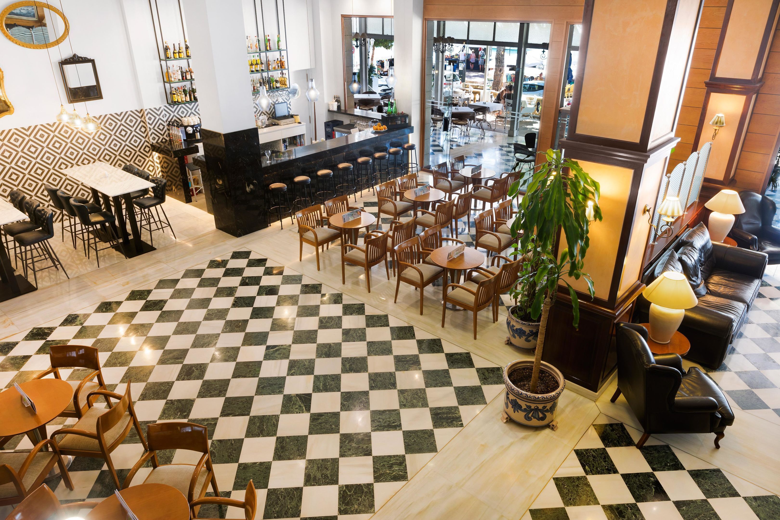Um lobby amplo com piso xadrez, diversas áreas de estar, um bar e uma grande planta em vaso.