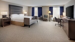 Quarto de hotel com uma cama grande, mesas de cabeceira com abajures, uma escrivaninha com cadeira, uma televisão e uma pequena mesa com uma cadeira.
